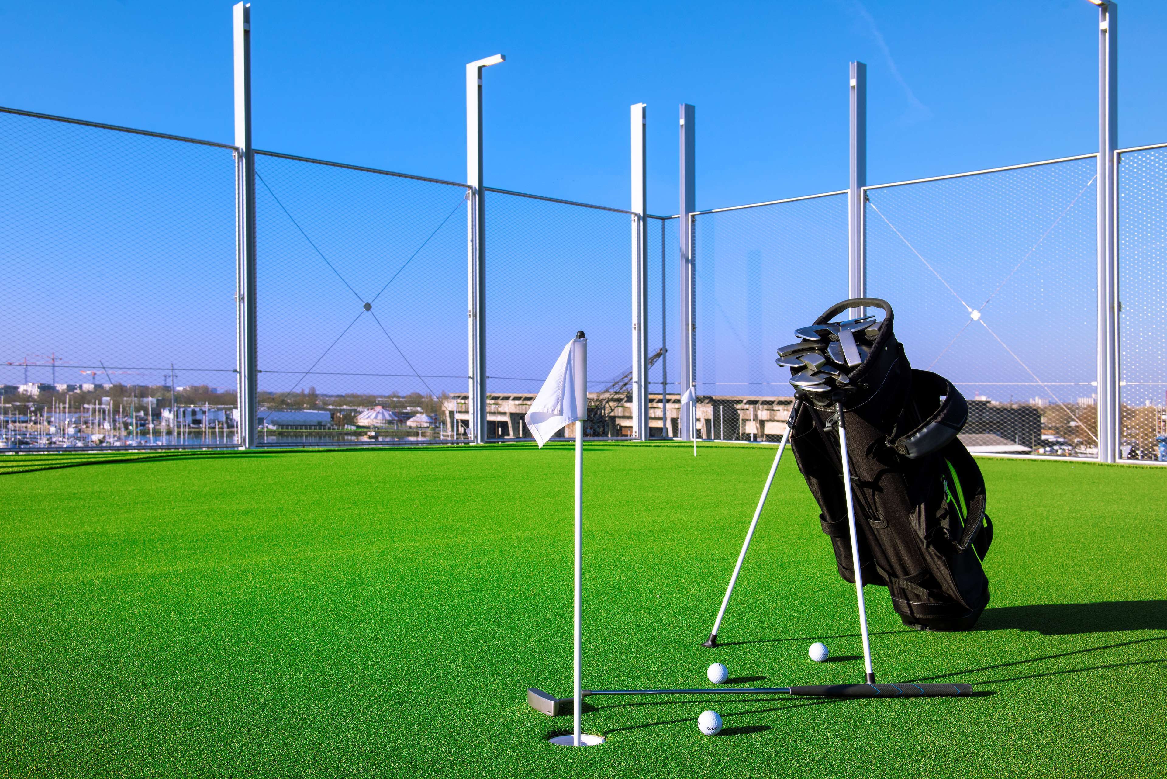 Uma área de treino de golfe com um campo verde, uma bolsa de golfe e um mastro com bandeira.