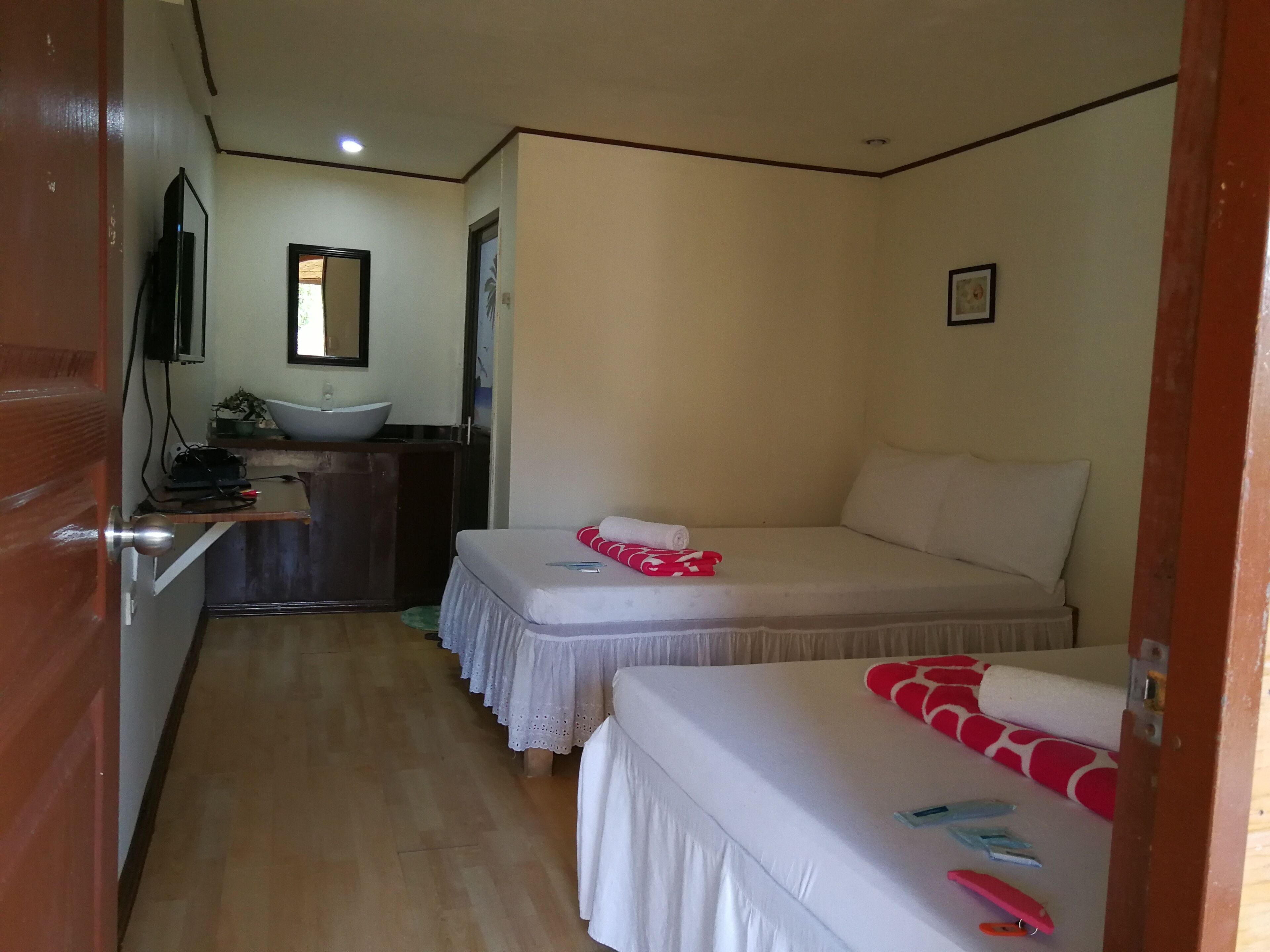 Um quarto de hotel pequeno com porta de madeira, cama com roupa de cama branca, uma pequena escrivaninha com pia e uma televisão fixada na parede.