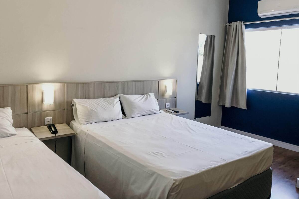 Quarto de hotel com duas camas, uma mesa de cabeceira com telefone, ar condicionado fixado na parede e janela com cortinas.
