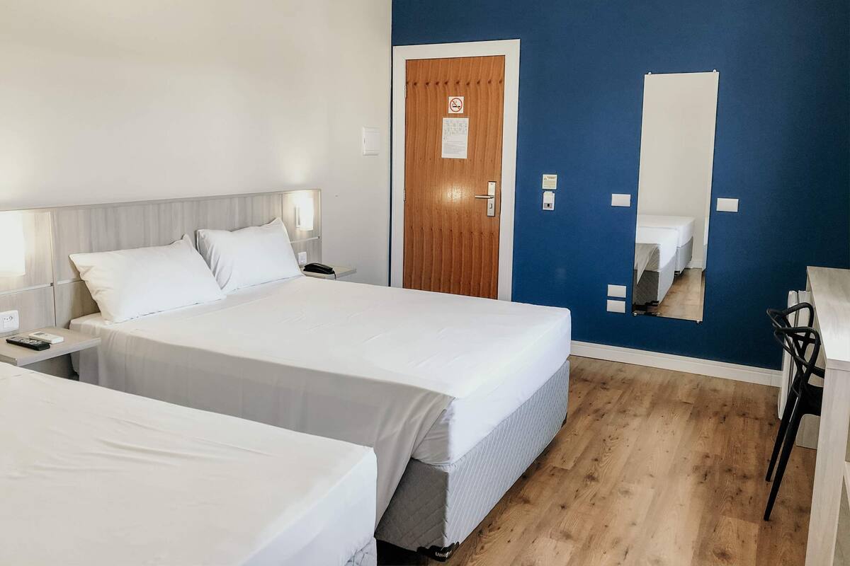 Quarto de hotel com duas camas, uma porta de madeira, um espelho e uma escrivaninha com uma cadeira.