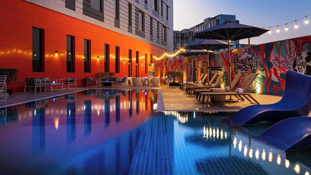 Piscina com espreguiçadeiras e guarda-sóis, cercada por um edifício com um mural colorido e luzes de festa.