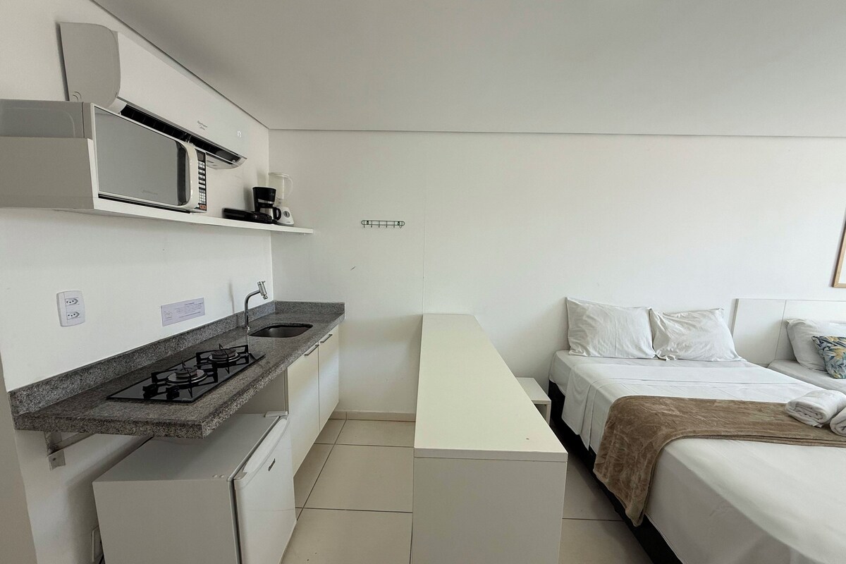 Quarto de hotel compacto com cozinha americana, cama com roupa de cama branca e uma pequena mesa de cabeceira.