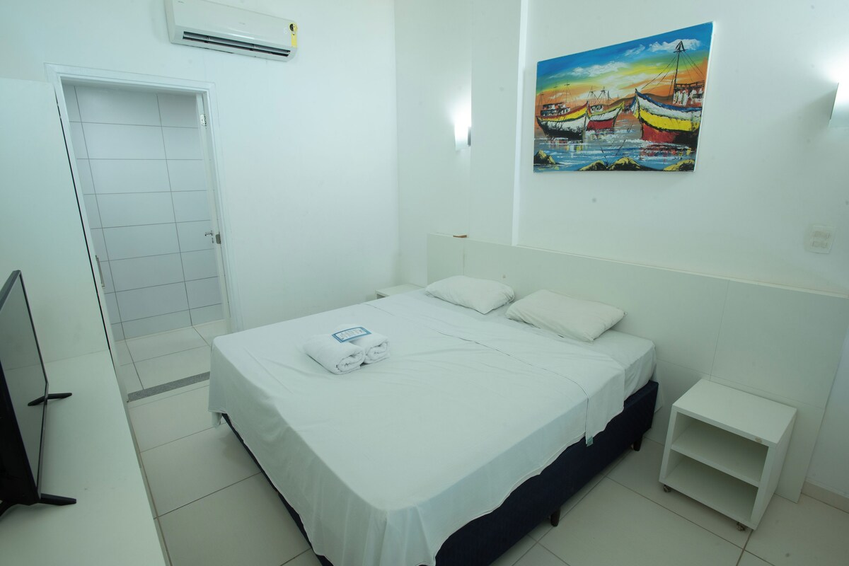 Quarto de hotel com cama, mesa de cabeceira, porta e um quadro na parede.