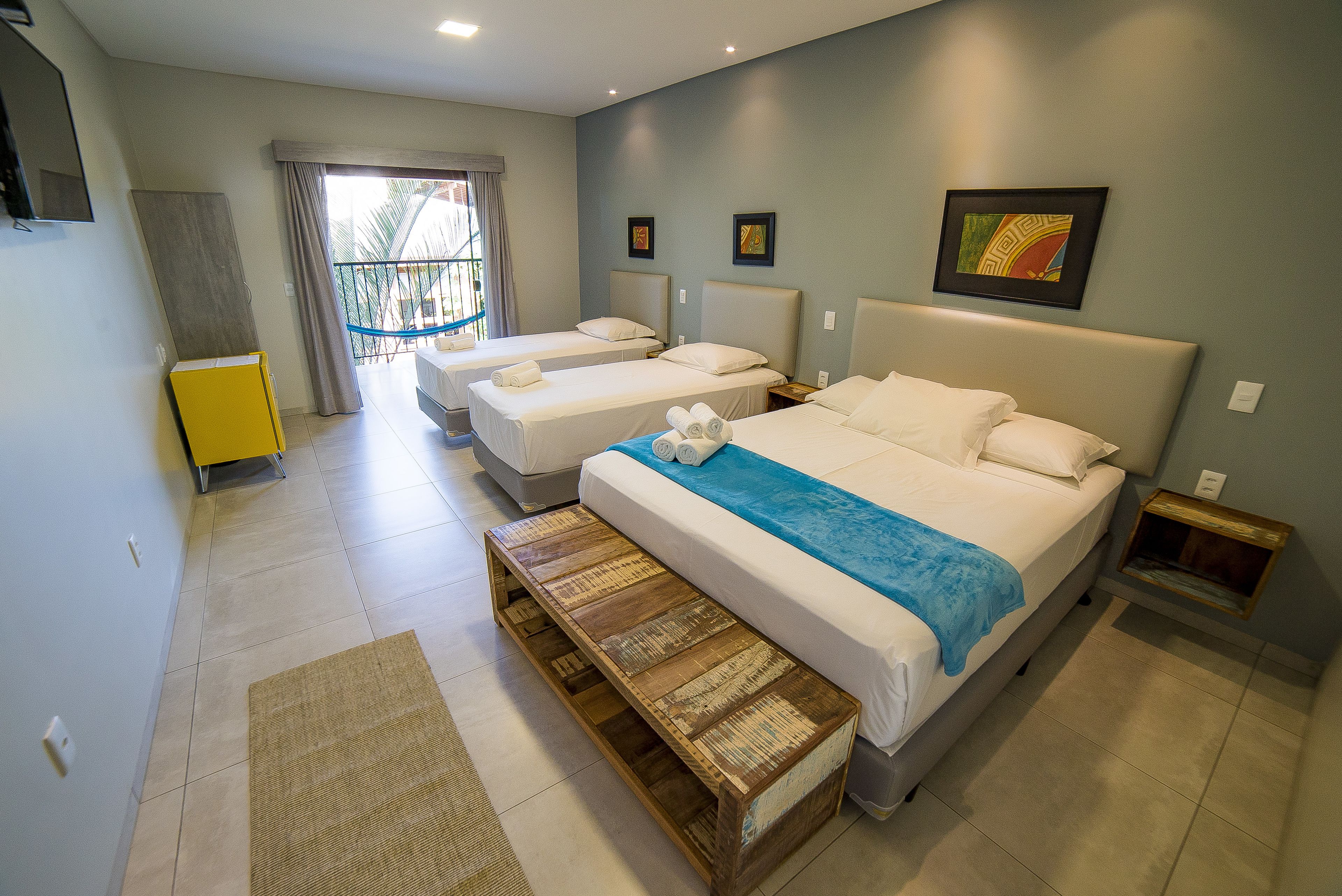 Quarto de hotel com duas camas de solteiro, uma cama grande, um banco de madeira, uma mesa pequena e uma varanda com vista.