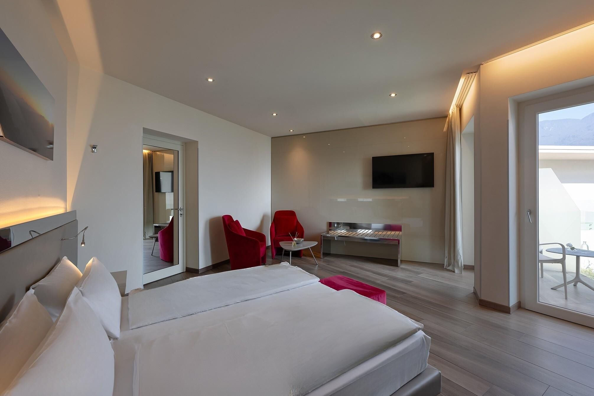 Quarto de hotel moderno com uma cama grande, uma TV fixada na parede e uma varanda com mesa e cadeiras.