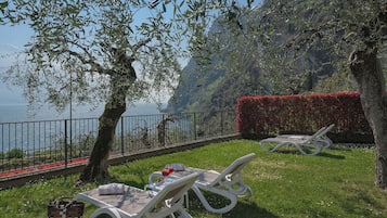 Um terraço com duas cadeiras de descanso brancas, uma mesa com uma bebida e vista para o mar e montanhas.