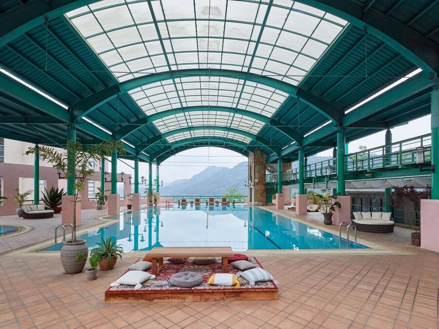 Piscina interior com teto de vidro, rodeada por plantas em vasos e áreas de estar.