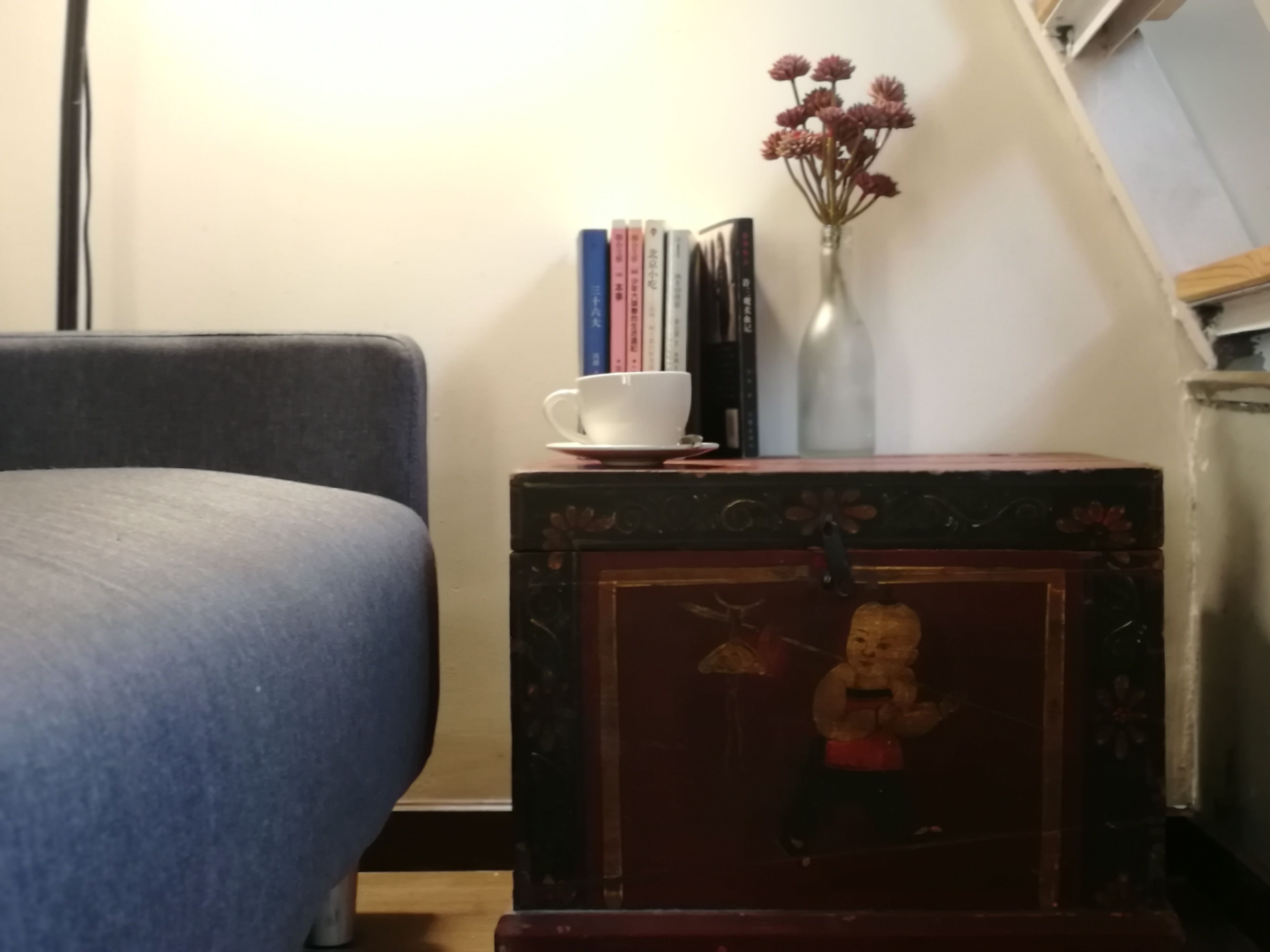 Quarto com uma poltrona azul, uma pequena mesa de cabeceira de madeira com uma xícara branca e livros, e um vaso com flores secas.