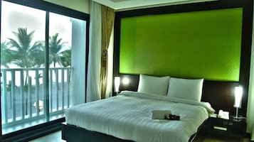 Quarto de hotel com uma cama grande, uma parede verde de destaque, uma varanda com vista para palmeiras e uma mesa de cabeceira com abajur.