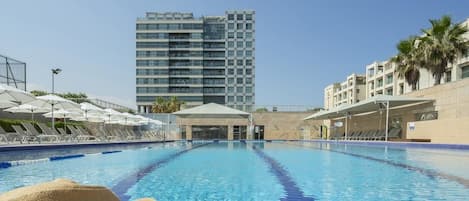 Piscina com espreguiçadeiras e guarda-sóis, um edifício alto ao fundo e um chapéu e óculos de natação à beira da piscina.