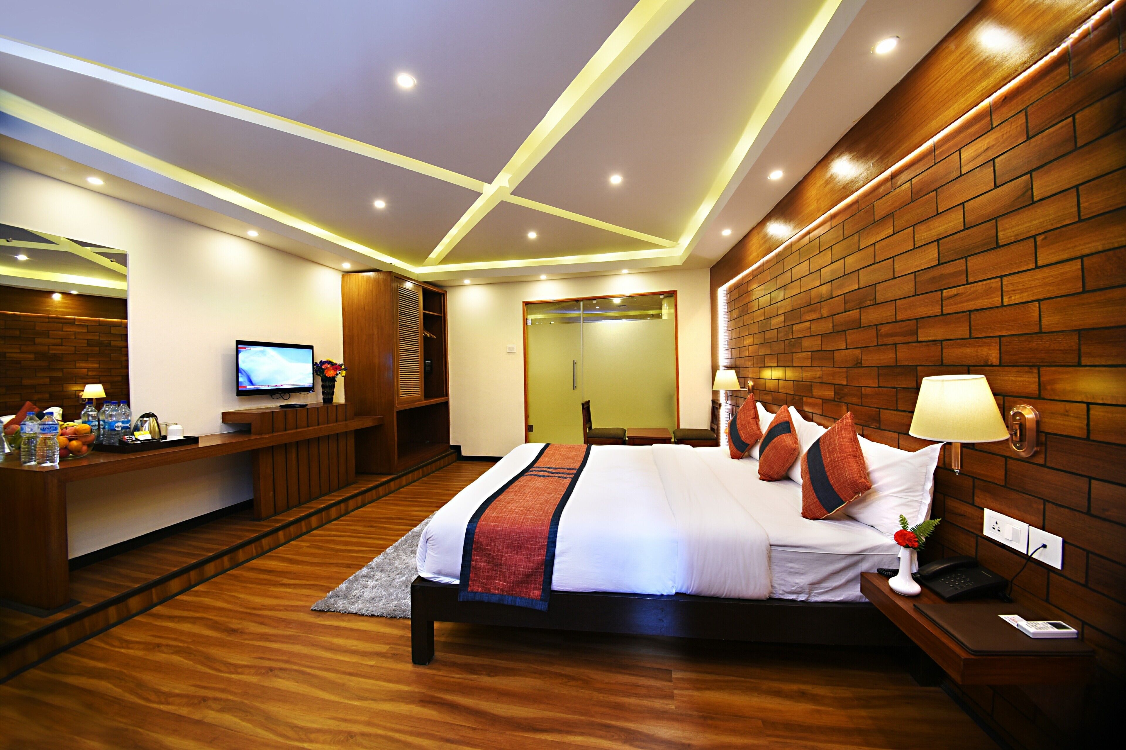 Quarto de hotel com piso de madeira, cama com roupa de cama branca e detalhes vermelhos, escrivaninha com computador, televisão e banheiro visível através de uma porta de vidro.