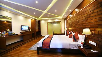 Quarto de hotel com piso de madeira, cama com roupa de cama branca e detalhes vermelhos, escrivaninha com computador, televisão e banheiro visível através de uma porta de vidro.