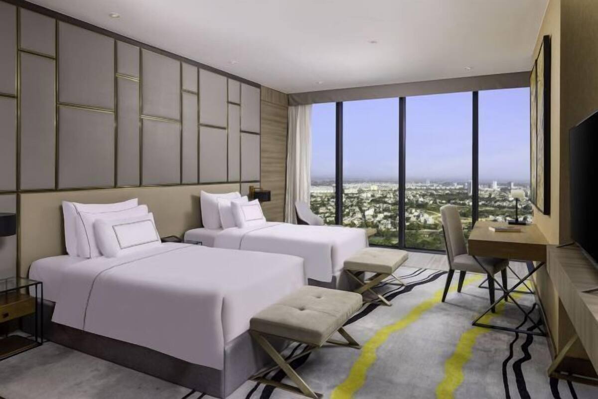 Quarto de hotel moderno com duas camas, uma escrivaninha e uma janela grande com vista para a cidade.