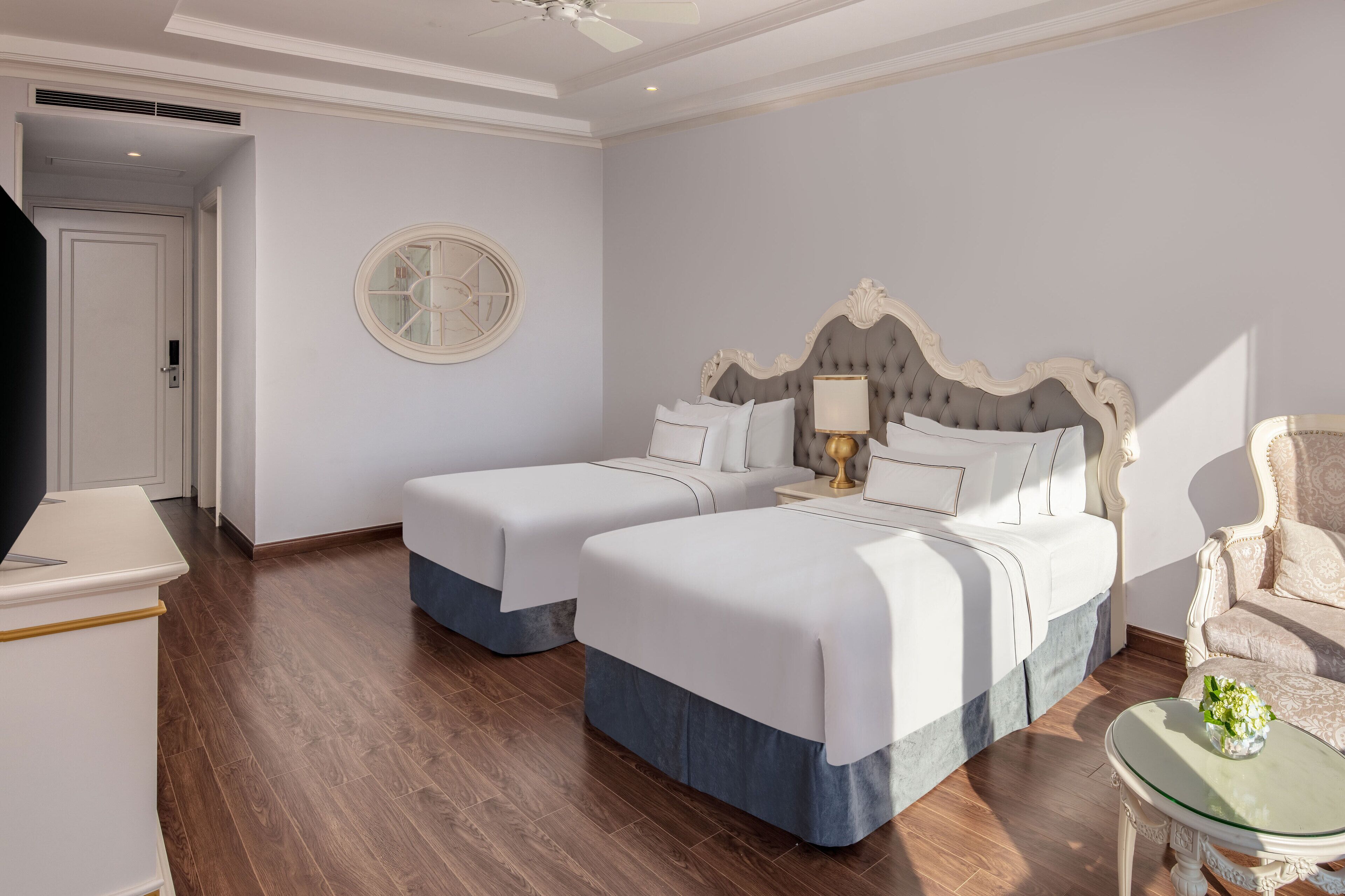 Quarto de hotel com duas camas, um relógio redondo na parede, um ventilador de teto e uma pequena mesa com um vaso.