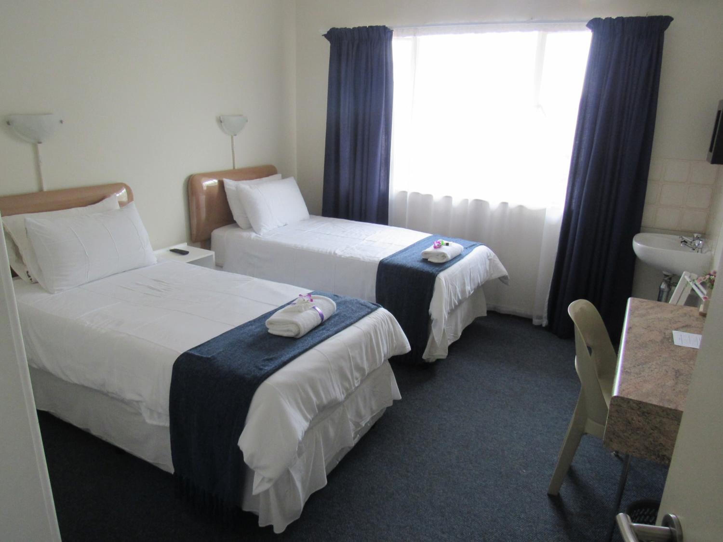 Quarto de hotel com duas camas, uma escrivaninha, uma cadeira, uma pia e uma janela com cortinas.