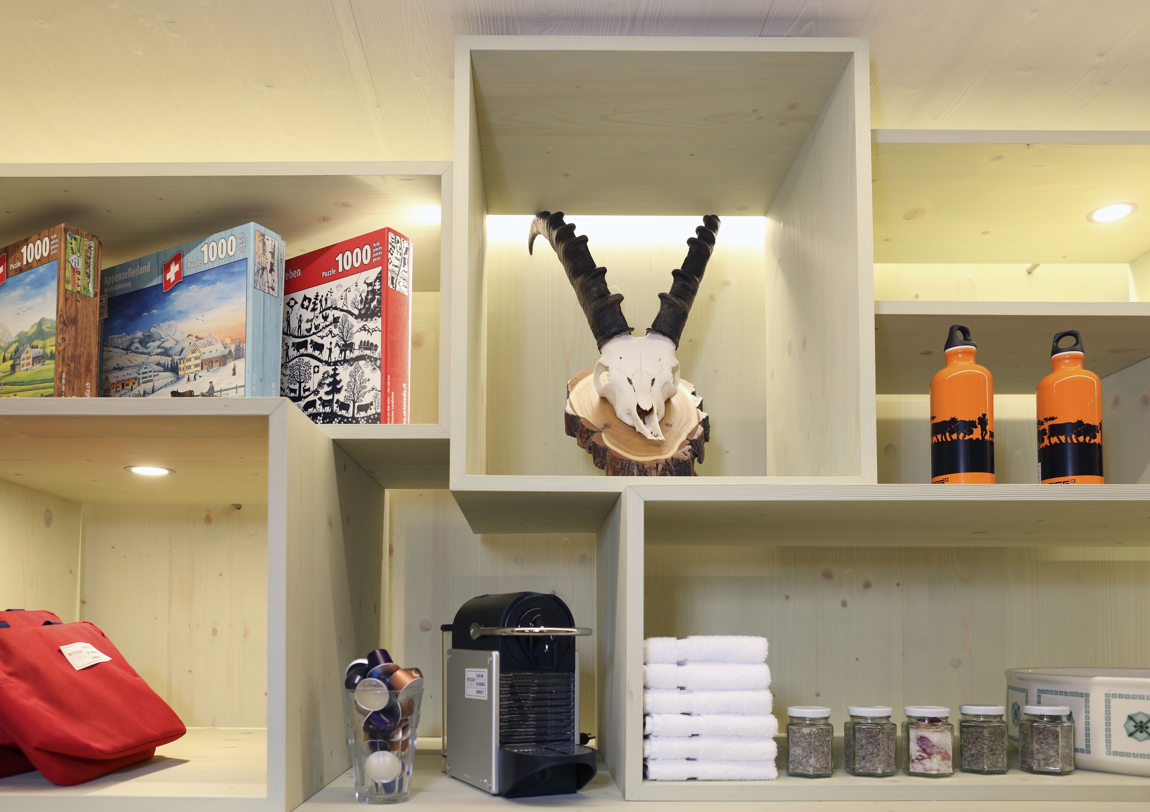 Uma estante com caixas de quebra-cabeça, uma decoração de caveira, garrafas de água, uma máquina de café, toalhas e potes.