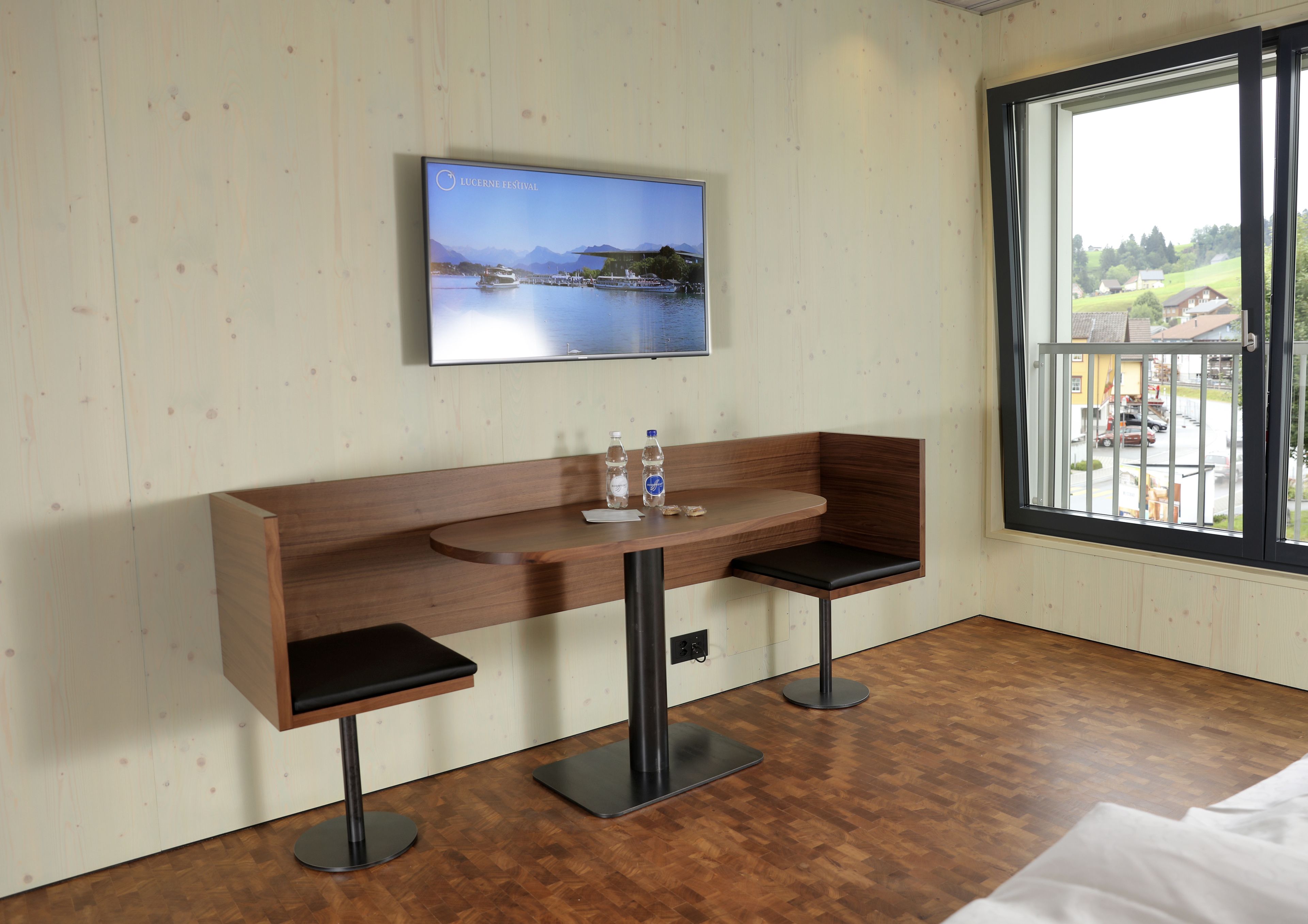 Quarto moderno com escrivaninha de madeira, duas cadeiras pretas, televisão exibindo uma vista panorâmica e janela com vista para um cais e barcos.