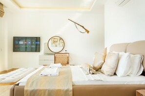 Quarto de hotel moderno com cama, sofá, televisão, espelho fixado na parede e um elemento decorativo.