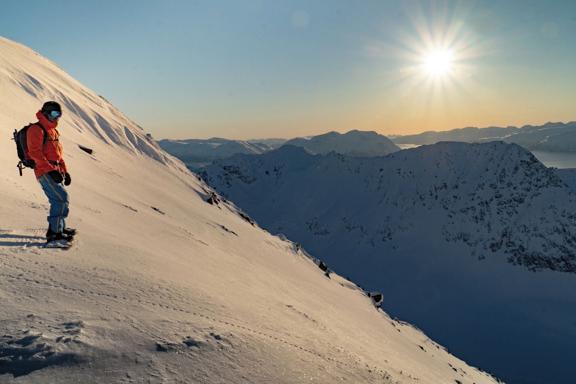 Uma pessoa praticando snowboard em uma encosta coberta de neve, com uma cordilheira ao fundo e o sol brilhando no céu.