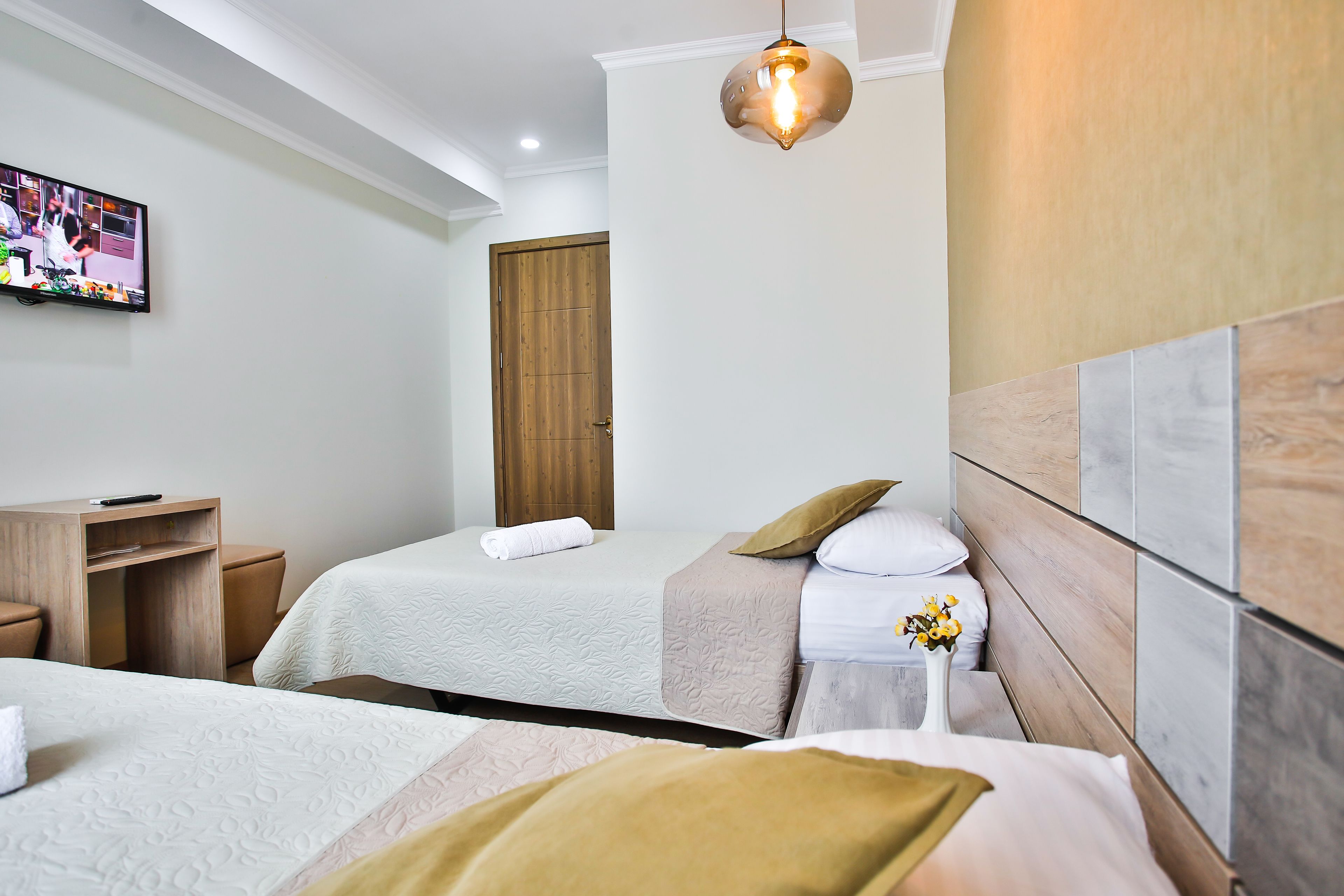 Quarto de hotel com duas camas, uma televisão, uma porta de madeira e uma pequena mesa com um vaso de flores.