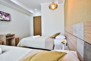 Quarto de hotel com duas camas, uma televisão, uma porta de madeira e uma pequena mesa com um vaso de flores.