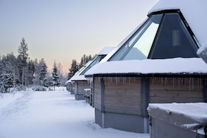 Fileira de cabanas de madeira com telhados cobertos de neve e pingentes de gelo, em meio a uma floresta nevada.