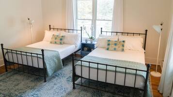 Quarto com duas camas de solteiro, uma mesinha de cabeceira, uma luminária e uma janela com cortinas.
