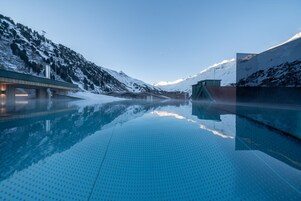 Piscina cristalina com montanhas ao fundo, uma ponte e um edifício com telhado inclinado.