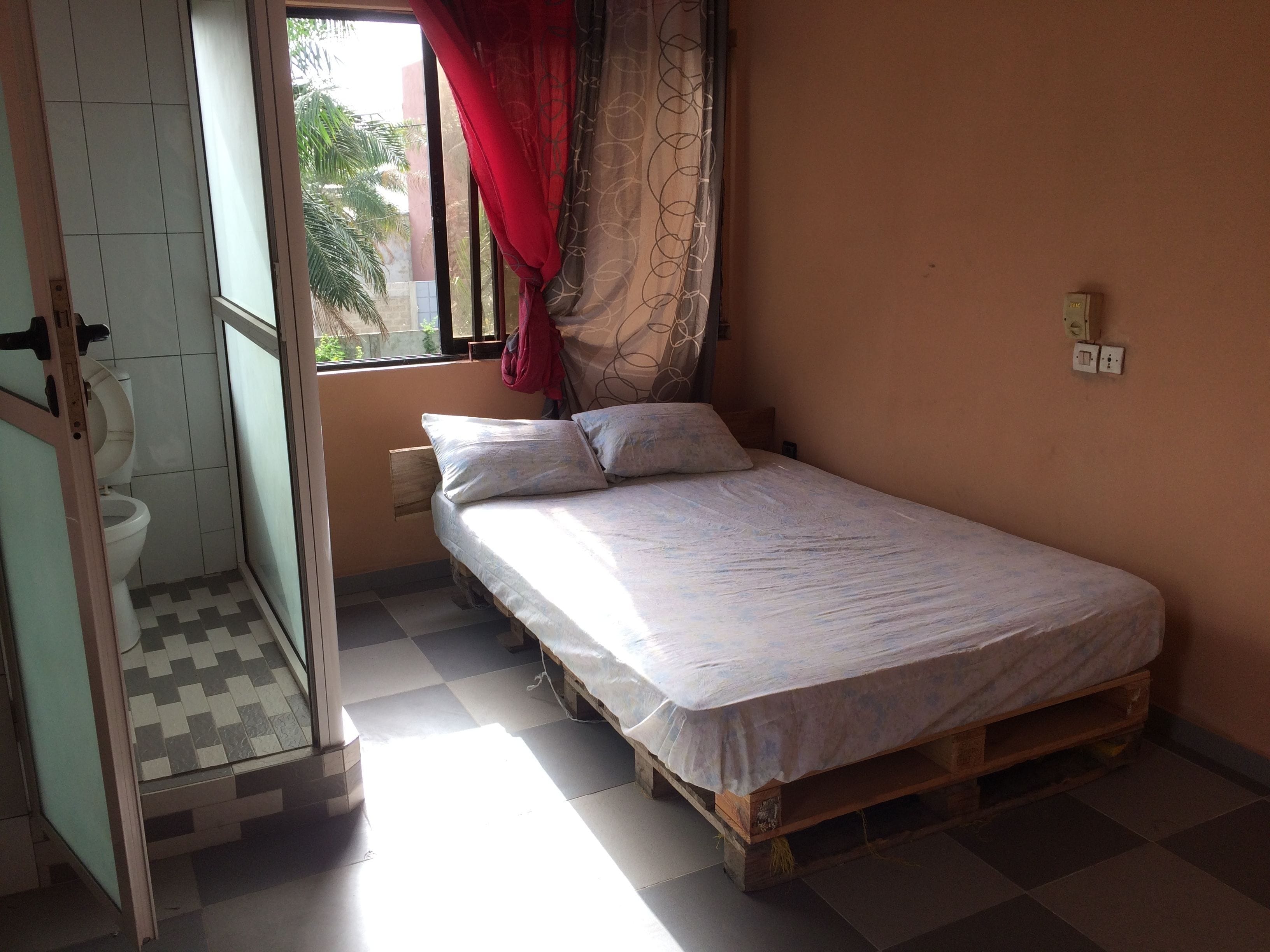 Quarto pequeno com uma cama de solteiro, estrutura de madeira, janela com cortinas vermelhas e vista para palmeiras.