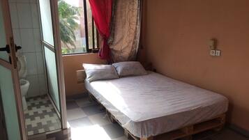 Quarto pequeno com uma cama de solteiro, estrutura de madeira, janela com cortinas vermelhas e vista para palmeiras.