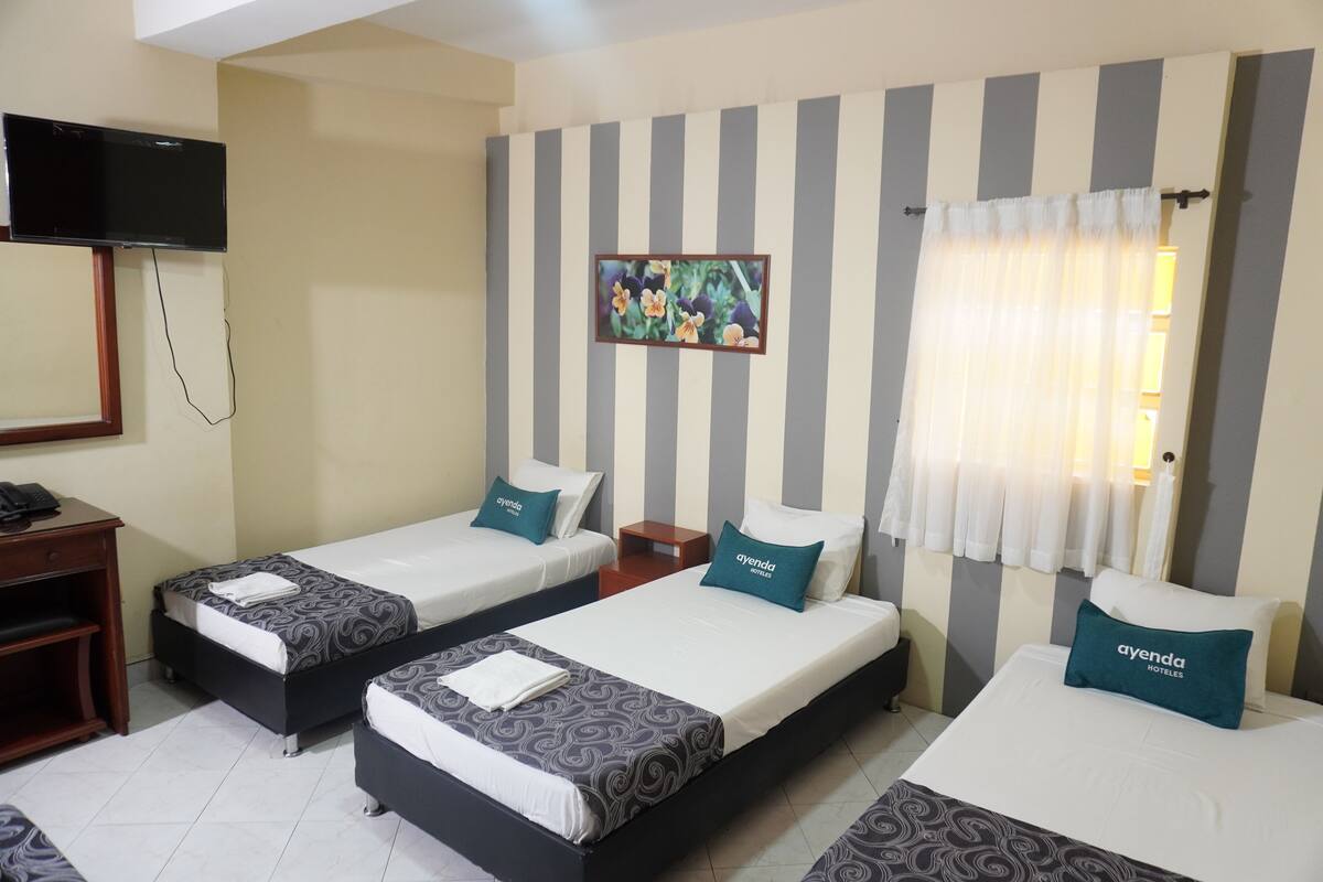 Quarto de hotel com três camas de solteiro, televisão de tela plana fixada na parede, uma mesa pequena e uma janela com cortinas.