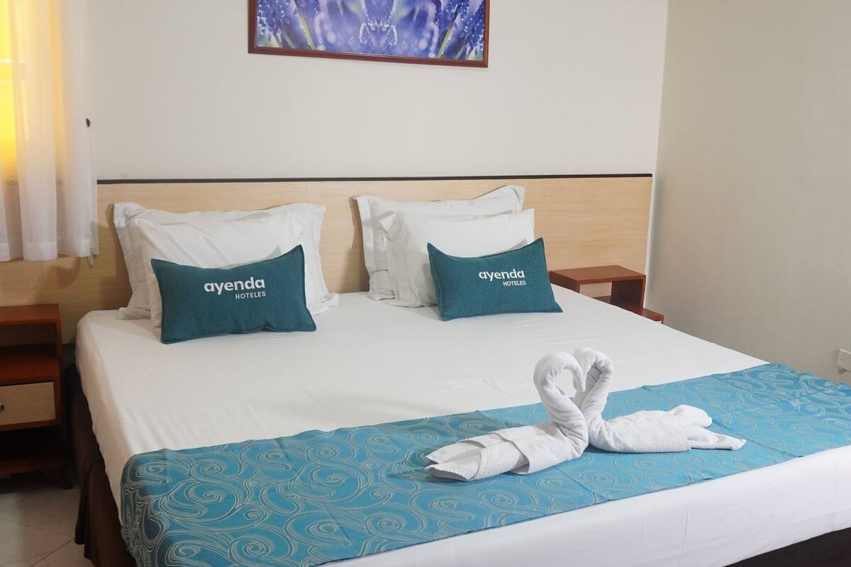 Quarto de hotel com uma cama bem arrumada, duas almofadas com o nome do hotel, uma obra de arte emoldurada na parede e uma mesa de cabeceira com uma luminária.