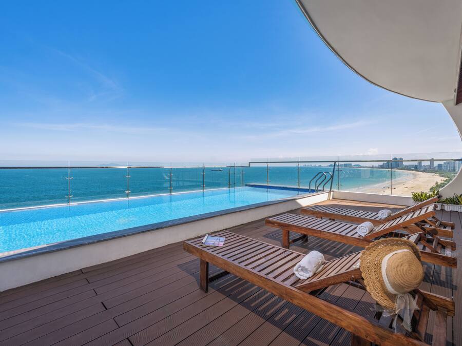 Um terraço com espreguiçadeiras de madeira, piscina e vista para a praia e oceano.