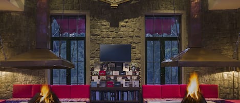 Um interior acolhedor com paredes de pedra, teto de madeira e lareira. Há estantes de livros e áreas de estar.