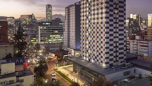 Um hotel moderno com fachada quadriculada, cercado por edifícios urbanos e uma rua movimentada ao anoitecer.