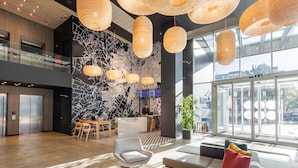 Um lobby moderno com pé-direito alto, luminárias pendentes grandes e feitas de fibra, balcão de recepção, área de estar com sofás e poltronas, e uma grande janela que proporciona iluminação natural.