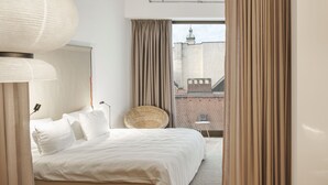 Quarto de hotel moderno com uma cama grande, uma mesa redonda pequena, uma cadeira de vime e vista para a paisagem urbana pela janela.