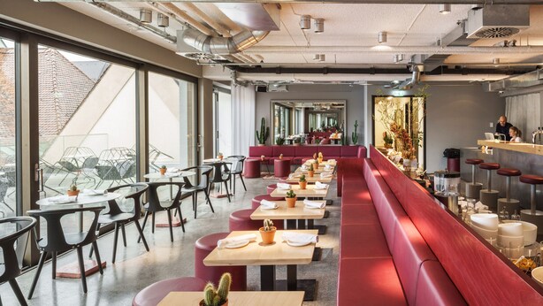 Restaurante moderno com um balcão comprido, assentos vermelhos e amplas janelas com vista para a paisagem urbana.