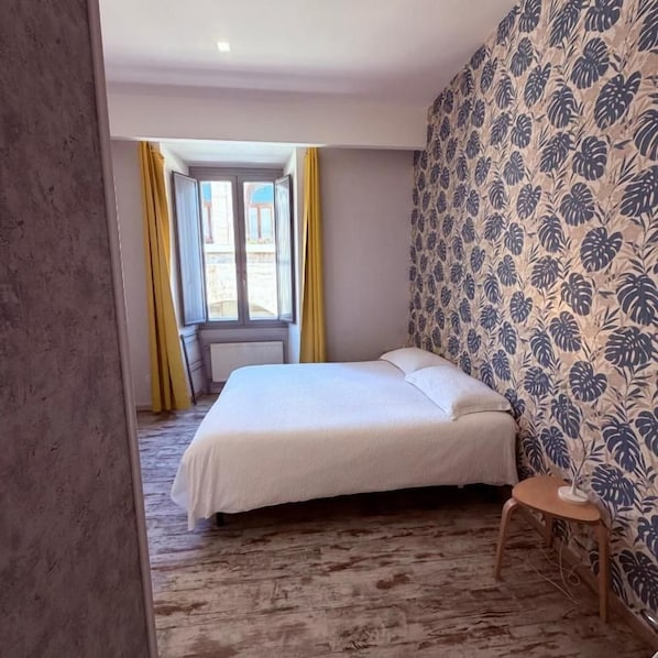 Quarto com cama, mesinha e janela com cortinas.