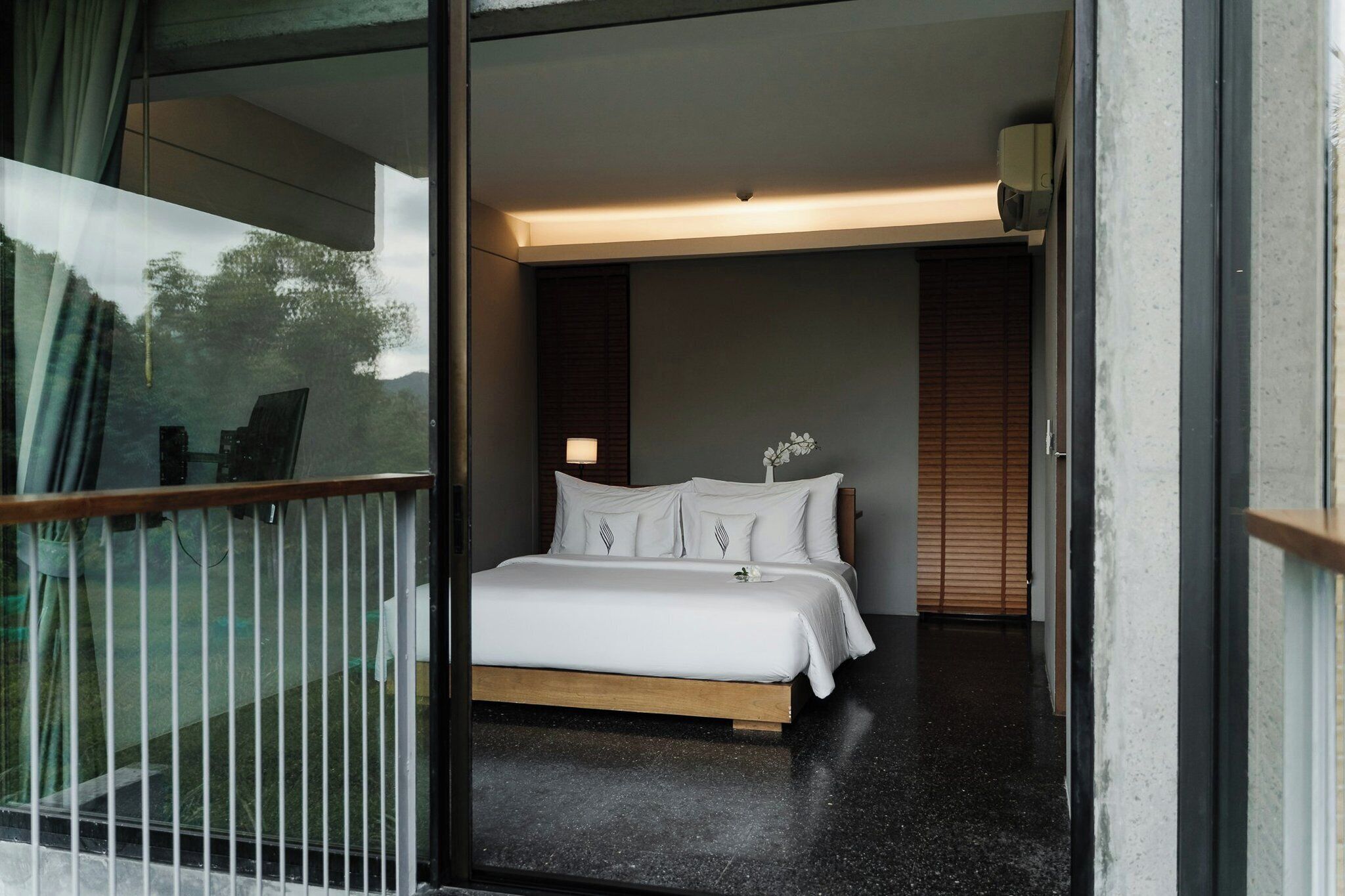 Quarto com cama, mesa de cabeceira, abajur e vista através de uma porta de vidro.