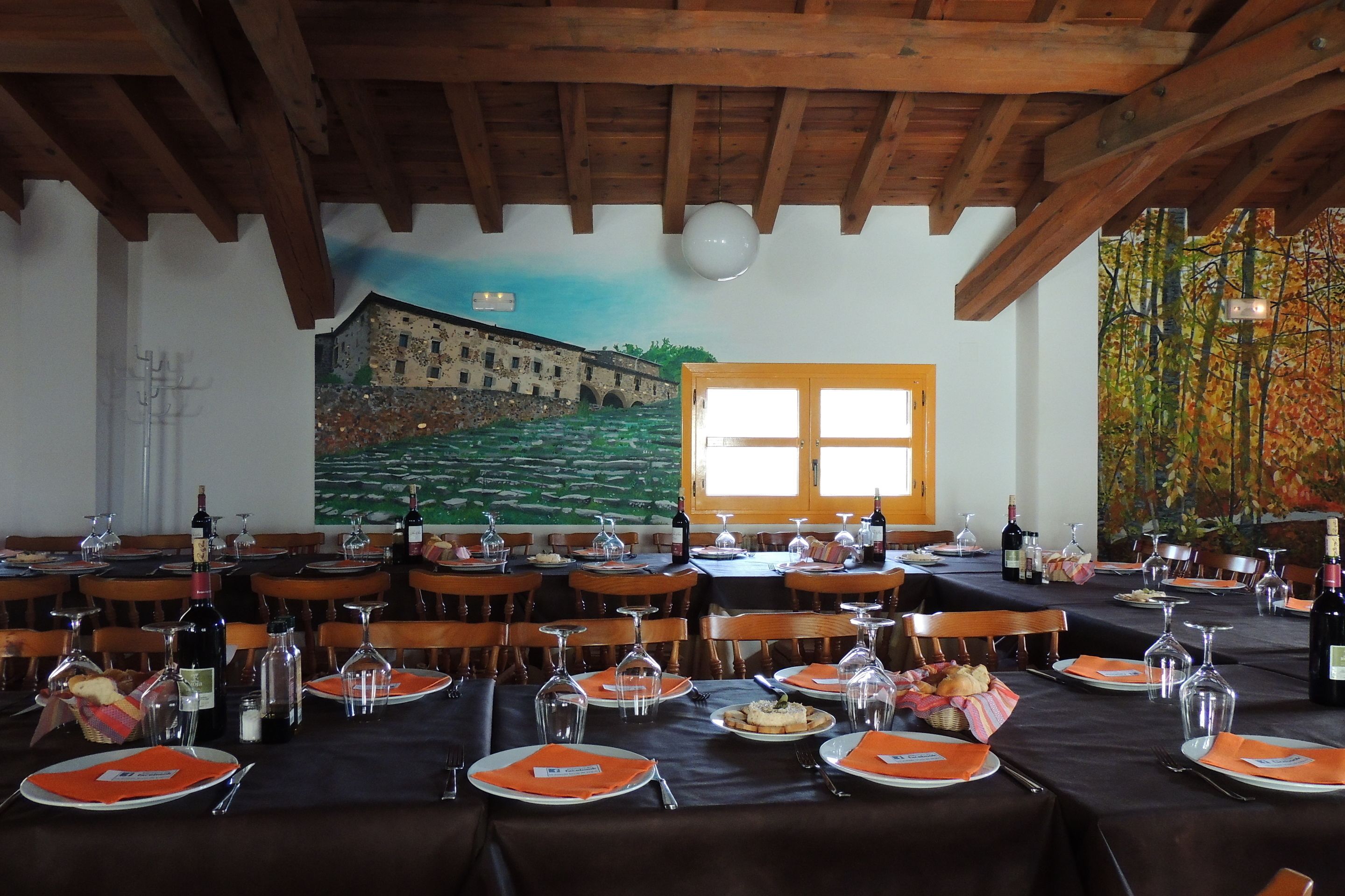 Uma sala de jantar com mesas dispostas para vários convidados, vigas de madeira no teto e um mural de um edifício na parede.