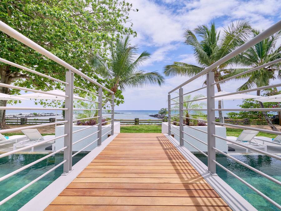 Um deck de madeira leva a uma piscina com guarda-corpos brancos e espreguiçadeiras, com vista para uma praia e o oceano.