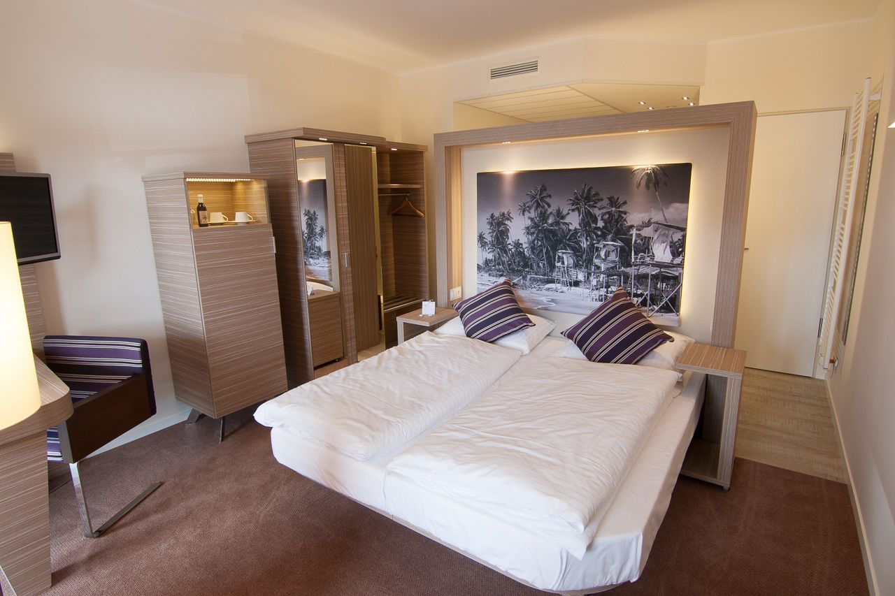 Quarto de hotel moderno com uma cama grande, um guarda-roupa, mesa de cabeceira e televisão na parede.
