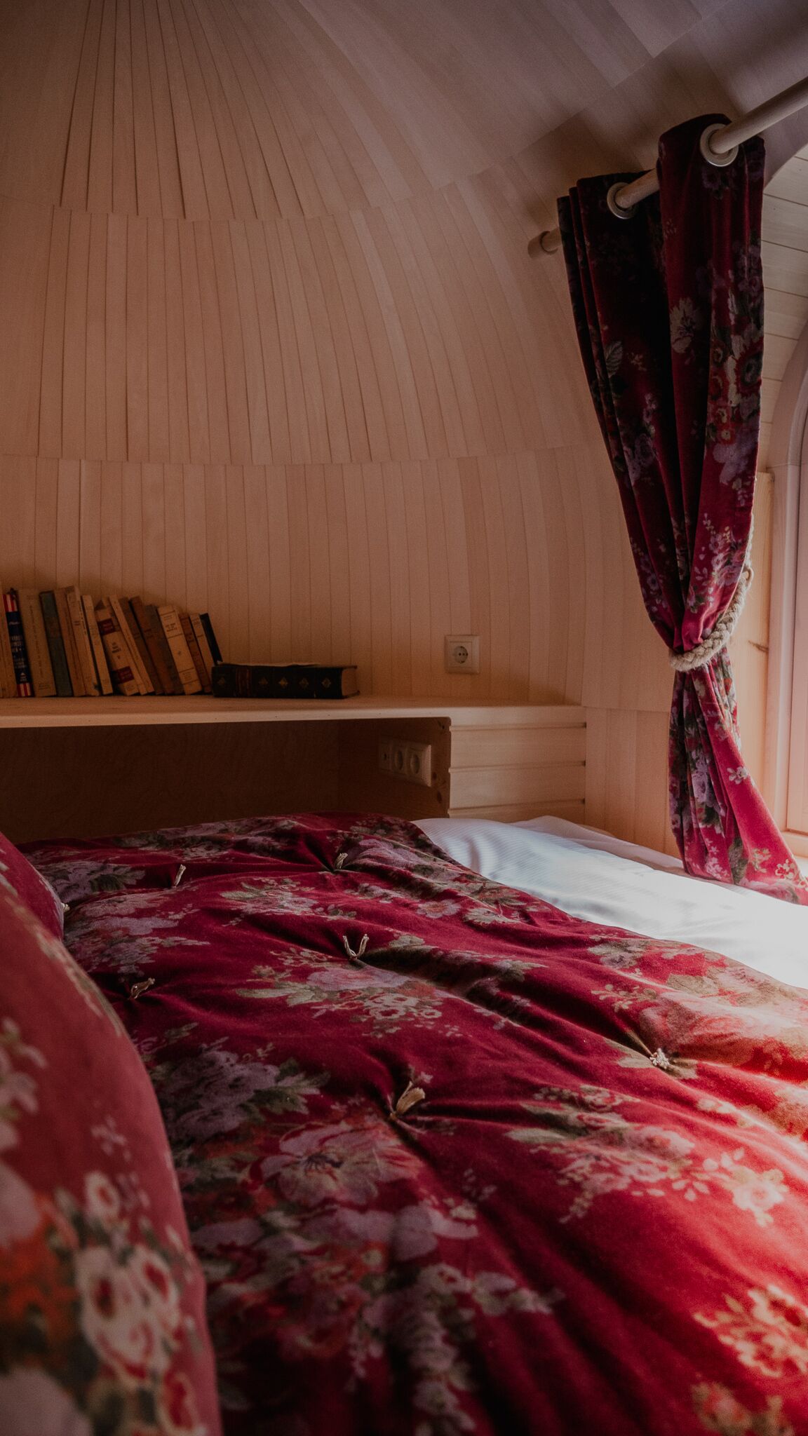 Um quarto com uma cama beliche de madeira, colcha vermelha florida e uma estante com livros.