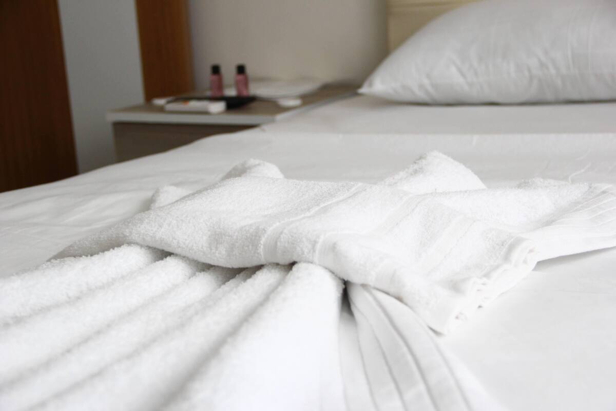 Cama bem arrumada com lençóis brancos e uma toalha dobrada.