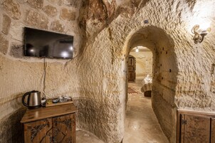 Um corredor com paredes de pedra, uma televisão fixada na parede, um armário de madeira com uma chaleira e uma pequena mesa com objetos sobre ela.