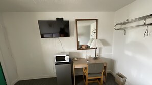 Quarto de hotel pequeno com cozinha compacta, escrivaninha, cadeira, televisão fixada na parede, espelho e luminária.