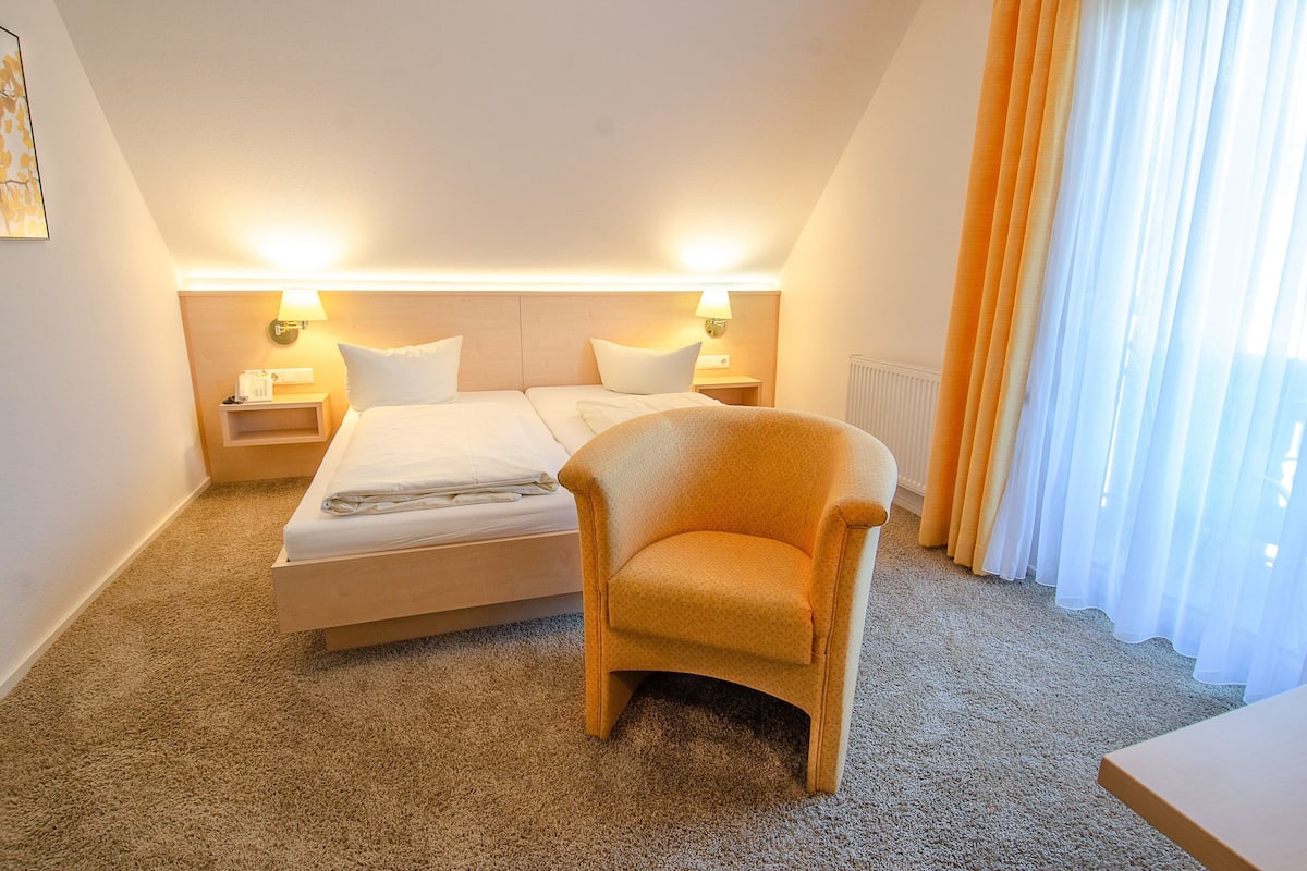 Quarto de hotel com uma cama, duas mesas de cabeceira, uma cadeira e uma janela com cortinas.