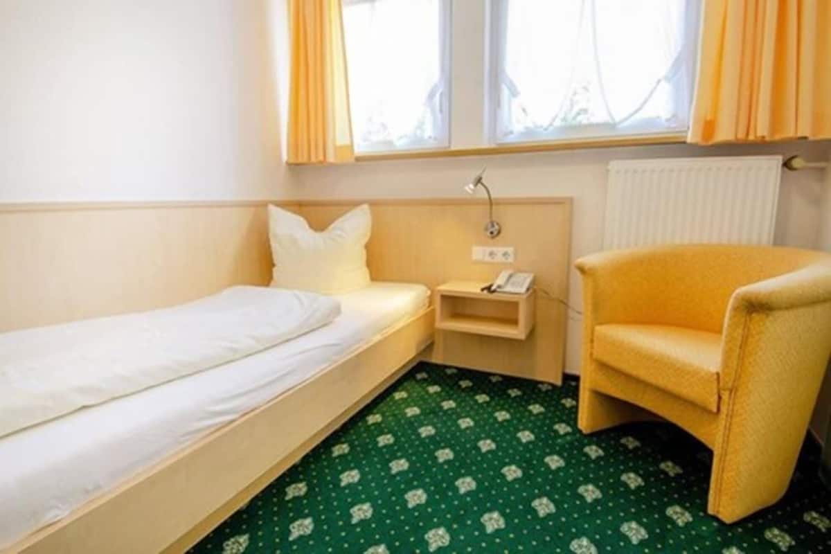 Quarto pequeno com uma cama de solteiro, piso com estampa verde, uma poltrona amarela e uma janela com cortinas.