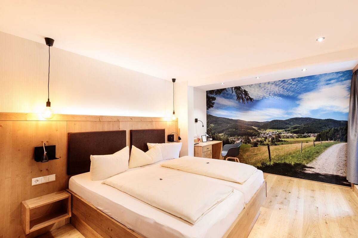Uma cama de casal com cabeceira de madeira, mesas de cabeceira, uma escrivaninha e vista para uma paisagem através de uma janela grande.